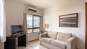 Quarto com televisão sobre uma escrivaninha, sofá, abajur e um quadro na parede.