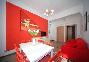 Uma área de refeições com mesa branca e cadeiras laranja, parede com detalhe vermelho e uma pintura, e sofá vermelho ao fundo.