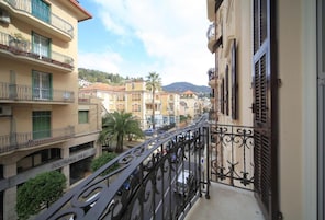 Varanda com grades de ferro forjado, vista para uma rua com edifícios e uma montanha ao fundo.