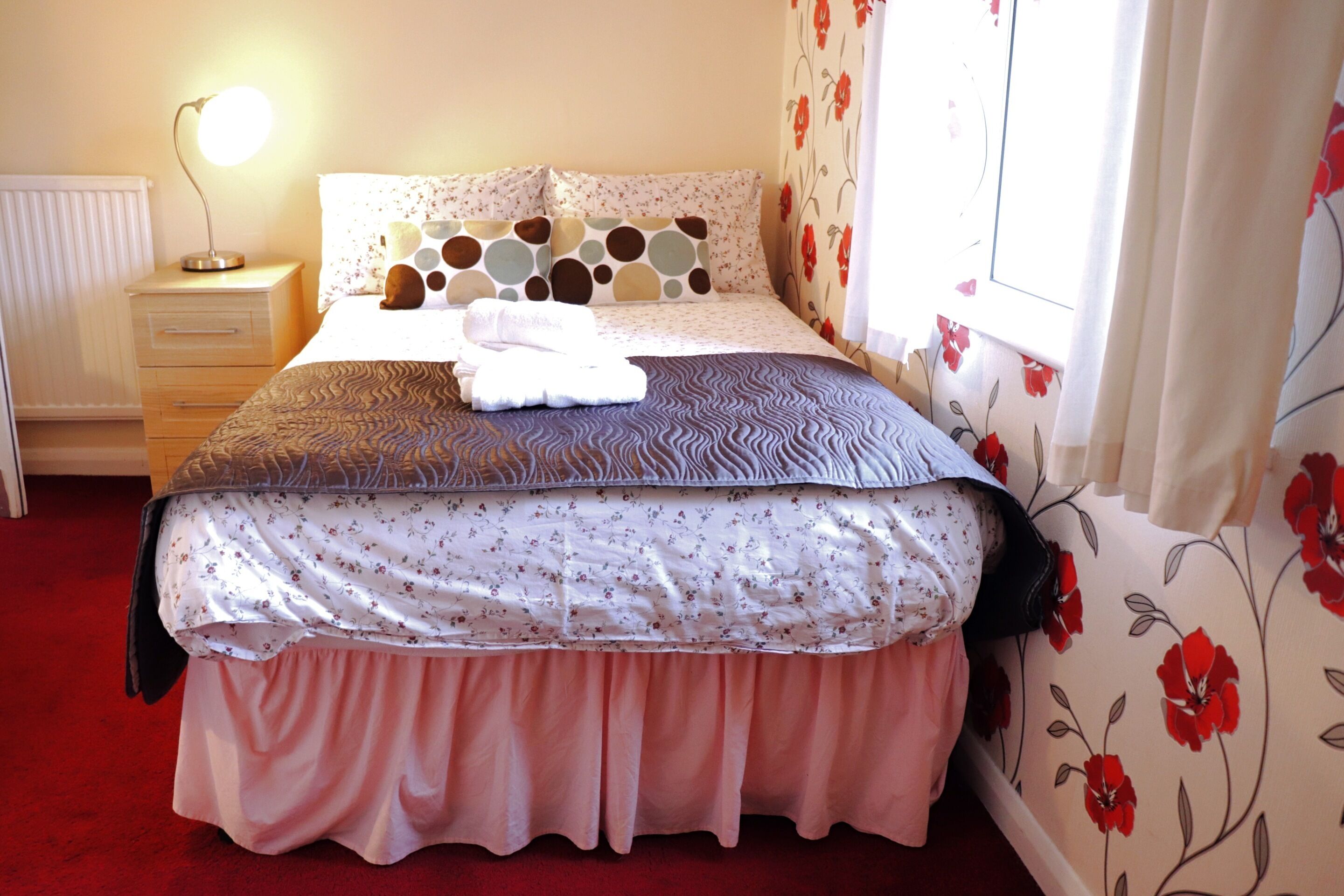 Cama bem arrumada com colcha estampada, luminária de cabeceira e papel de parede floral.