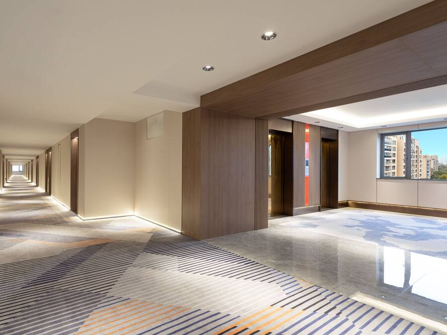 Corredor moderno de hotel com carpete estampado, painéis de madeira e vista para a cidade através de grandes janelas.