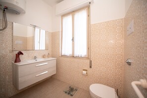 Casa de banho com lavatório branco, espelho e sanita, paredes estampadas e janela com cortinas.