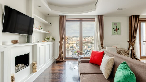 Uma sala de estar moderna com televisão de tela plana, sofá com almofadas, lareira e varanda com vista para a cidade.