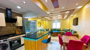 Uma cozinha moderna com uma ilha central, uma área de jantar com mesa de madeira e cadeiras coloridas, e um espaço de estar acolhedor com almofadas estampadas.