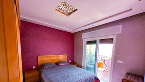 Um quarto com cama de madeira, mesa de cabeceira, um pequeno banquinho de madeira, uma porta de vidro deslizante que dá para uma área externa e um lustre no teto.