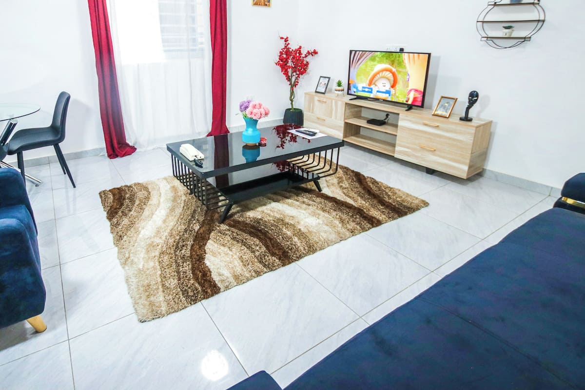 Uma sala de estar moderna com uma mesa de centro de vidro, cortinas vermelhas, uma TV de tela plana, um relógio e um armário de madeira.