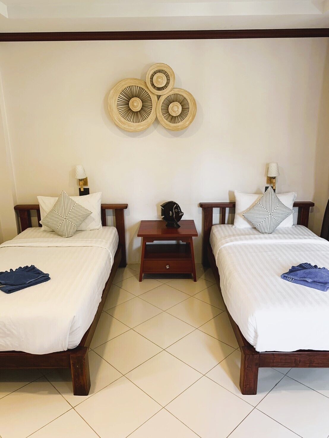 Quarto de hotel com duas camas, cada uma com estrutura de madeira e roupa de cama branca. Há peças de arte decorativa nas paredes acima das camas e uma pequena mesa de cabeceira de madeira entre elas.