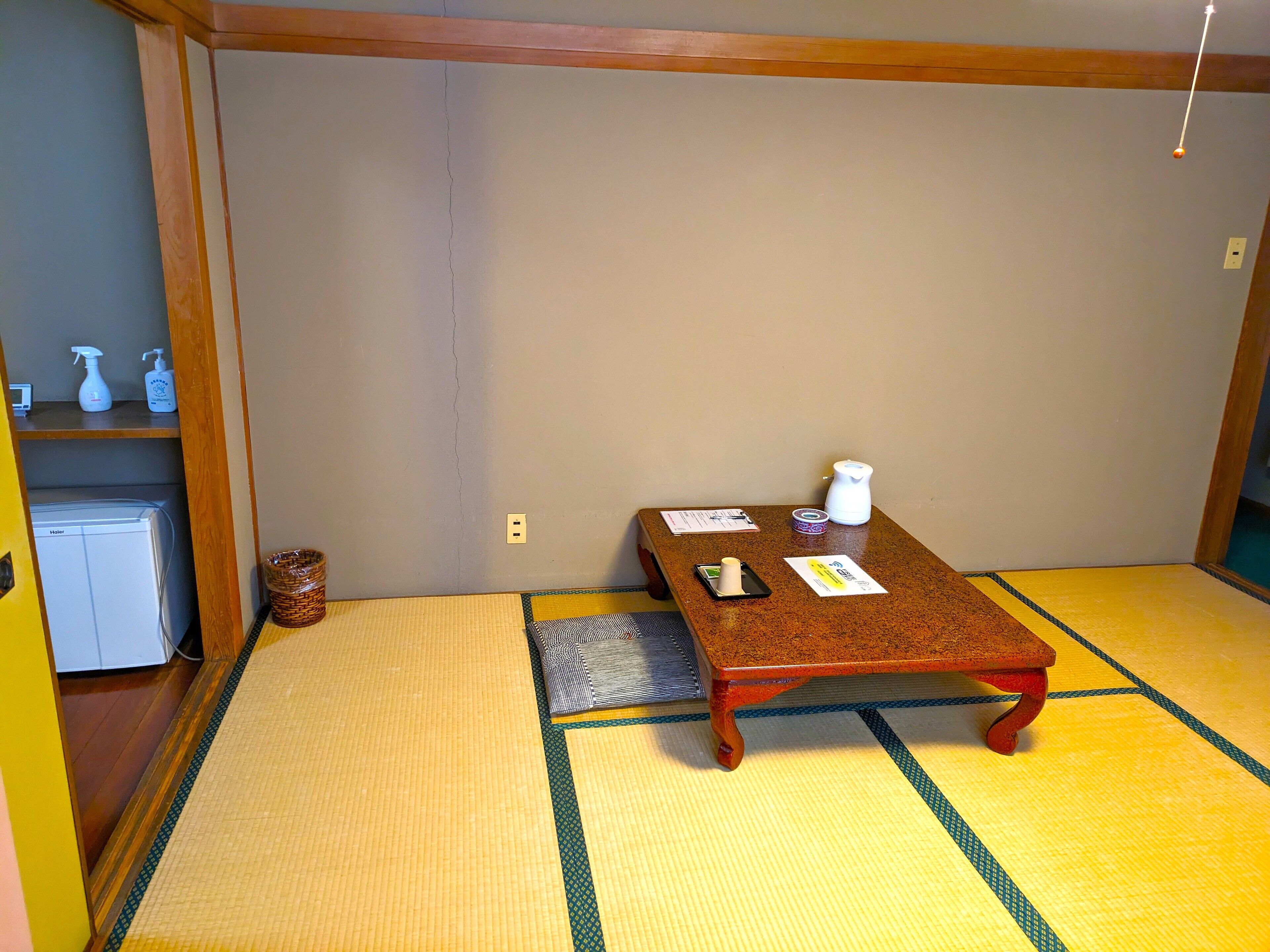 Quarto com piso de esteira de tatame, uma mesa de madeira com um jarro branco e um tabuleiro preto, e um pequeno aquecedor branco à esquerda.