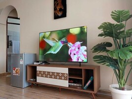 Sala de estar com televisão de tela plana em suporte de madeira, geladeira, planta em vaso e pintura pendurada na parede.