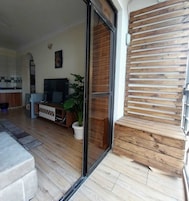 Uma sala de estar moderna com uma porta de vidro deslizante que dá para uma varanda, uma televisão, uma planta e uma área de cozinha ao fundo.