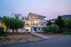 Um edifício moderno de dois andares chamado Lekka's House, com um caminho de pedra que leva à entrada, rodeado por vegetação e um fundo de colinas.