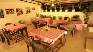Restaurante com várias mesas cobertas por toalhas quadriculadas vermelhas e brancas, rodeado por plantas em vasos e cadeiras.