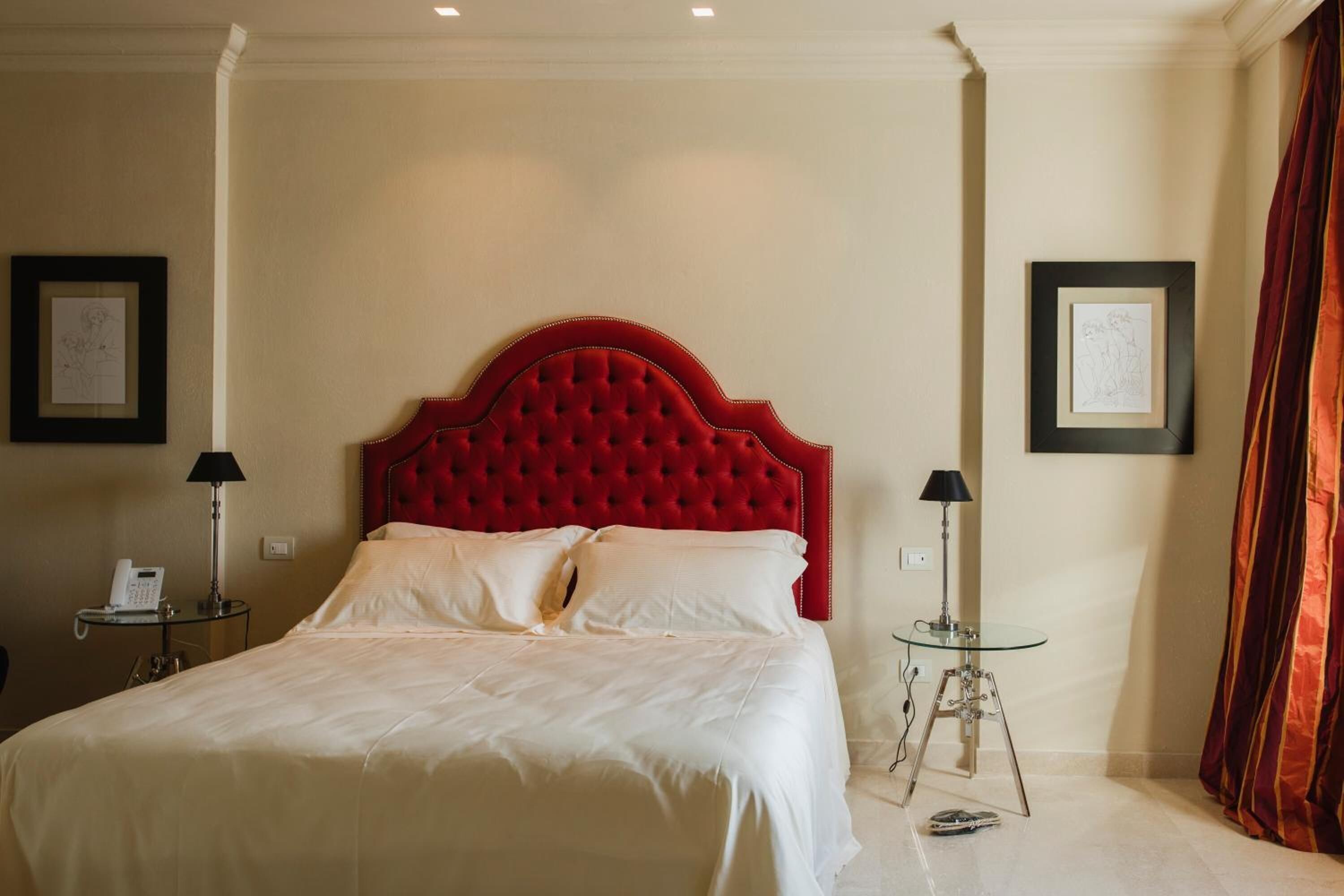 Quarto de hotel com cabeceira estofada vermelha, espalha branco, duas mesas de cabeceira com abajures e quadros nas paredes.