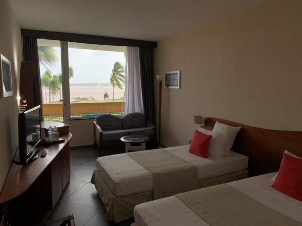 Quarto de hotel com cama, sofá, escrivaninha e vista para a praia.