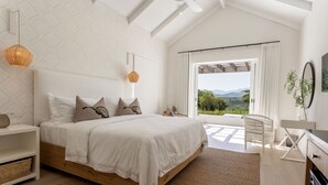 Quarto com cama grande, mesa de cabeceira, escrivaninha, cadeira e vista para o exterior.