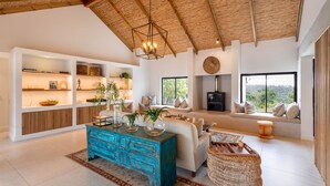 Uma sala de estar ampla, com teto alto, estantes embutidas, um aparador azul central, um sofá e uma janela grande com vista para o exterior.