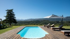 Piscina retangular com um deck de madeira, cercada por espreguiçadeiras e um grande guarda-sol.