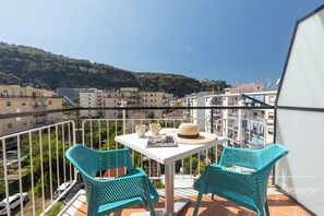 Uma varanda com duas cadeiras turquesa, uma mesa com chapéu e uma revista, e vista para uma cidade no alto de uma colina.
