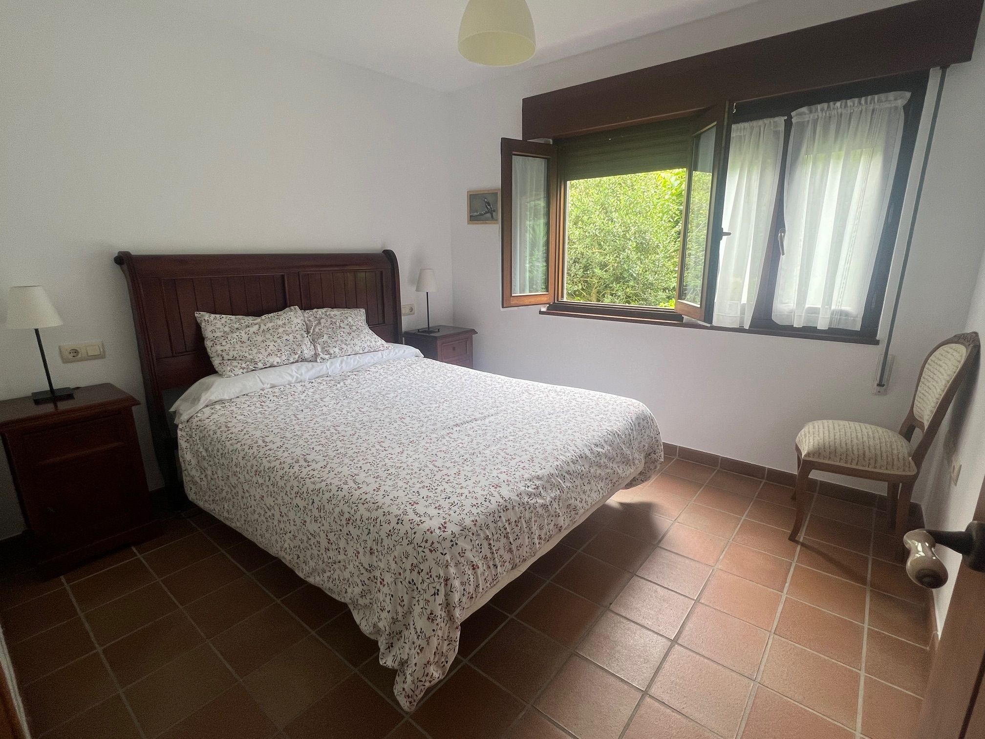 Quarto com cama, mesa de cabeceira, cadeira, janela com cortinas e vista para o verde lá fora.