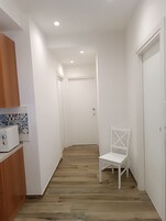 Um corredor estreito com paredes brancas, soalho de madeira e uma cadeira branca. Há armários e um micro-ondas à esquerda, e uma porta no final.
