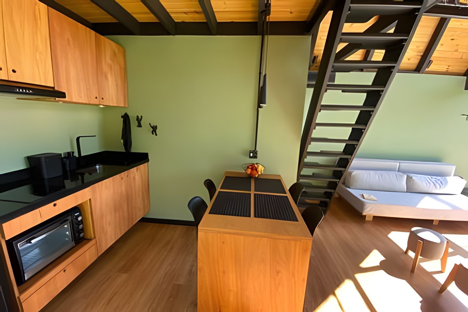 Cozinha compacta com armários de madeira, forno embutido e área de refeições com mesa e cadeiras. É visível um mezanino com uma cama e uma escada.