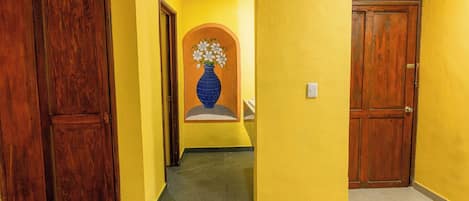 Um corredor de hotel com portas de madeira, parede amarela e um vaso azul com flores sobre um pedestal.