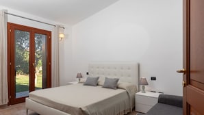 Quarto com cama grande, mesa de cabeceira, uma cadeira e porta de vidro deslizante que dá para um jardim.