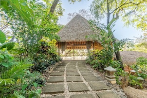Uma cabana com telhado de palha, com um caminho de pedra que a leva até ela, cercada por vegetação exuberante.