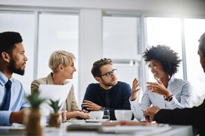 Um grupo de pessoas em uma sala de reunião com uma janela grande, uma mesa com documentos e uma xícara.
