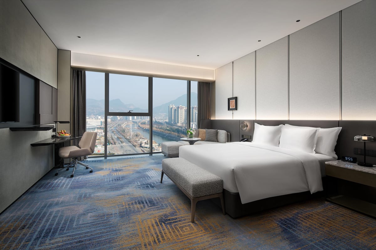 Quarto de hotel moderno com uma cama grande, uma escrivaninha, uma cadeira e vista para o horizonte urbano através de amplas janelas.