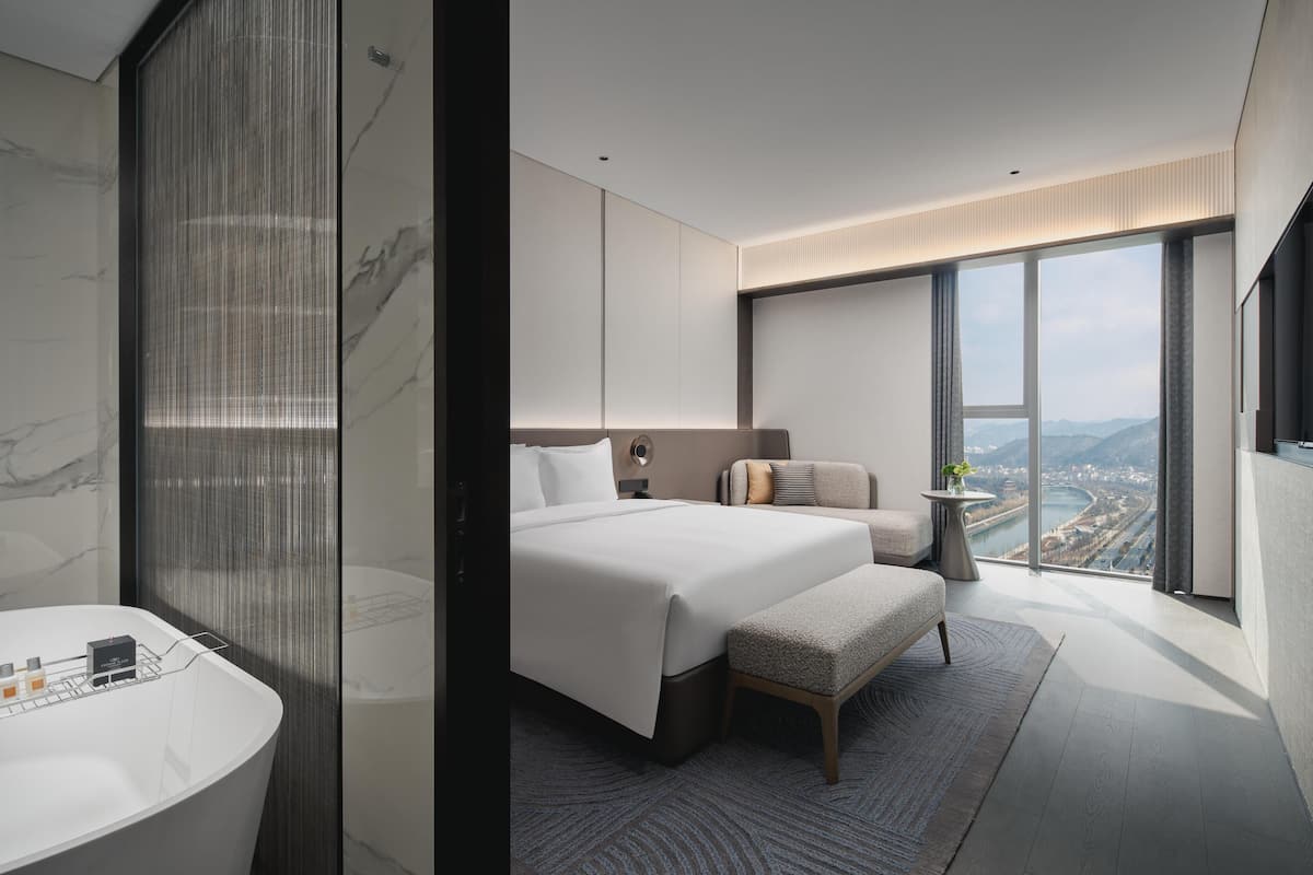 Quarto de hotel moderno com uma cama grande, área de estar, vista para a cidade e banheiro com banheira espaçosa.