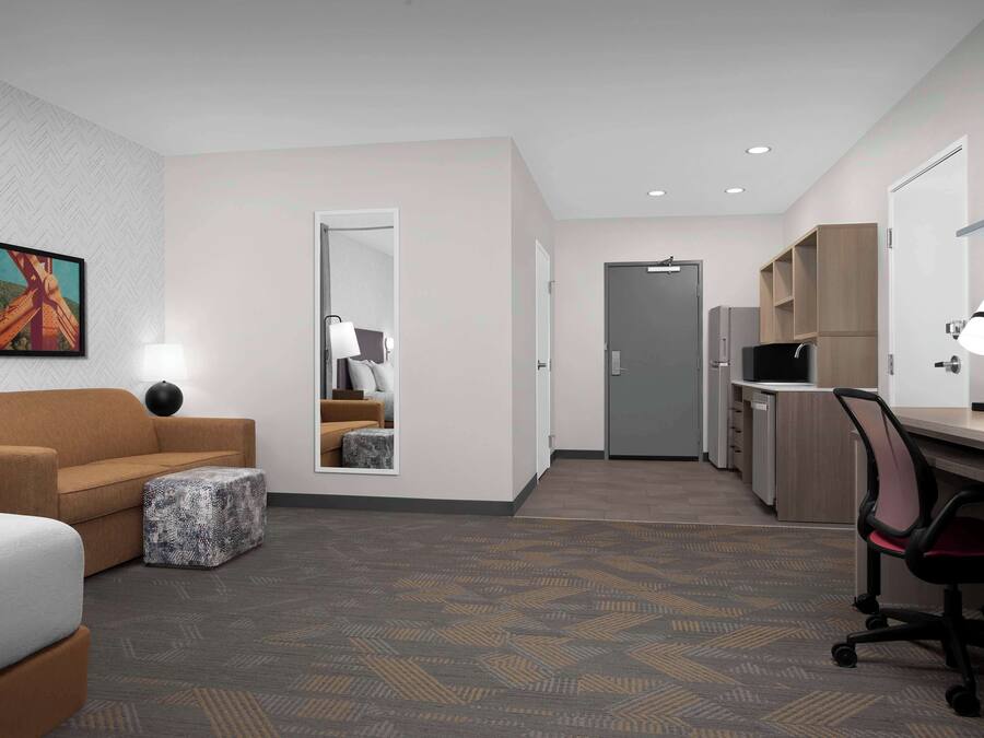 Quarto de hotel moderno com sofá, um pequeno banquinho, escrivaninha com cadeira, espelho e área de cozinha compacta.