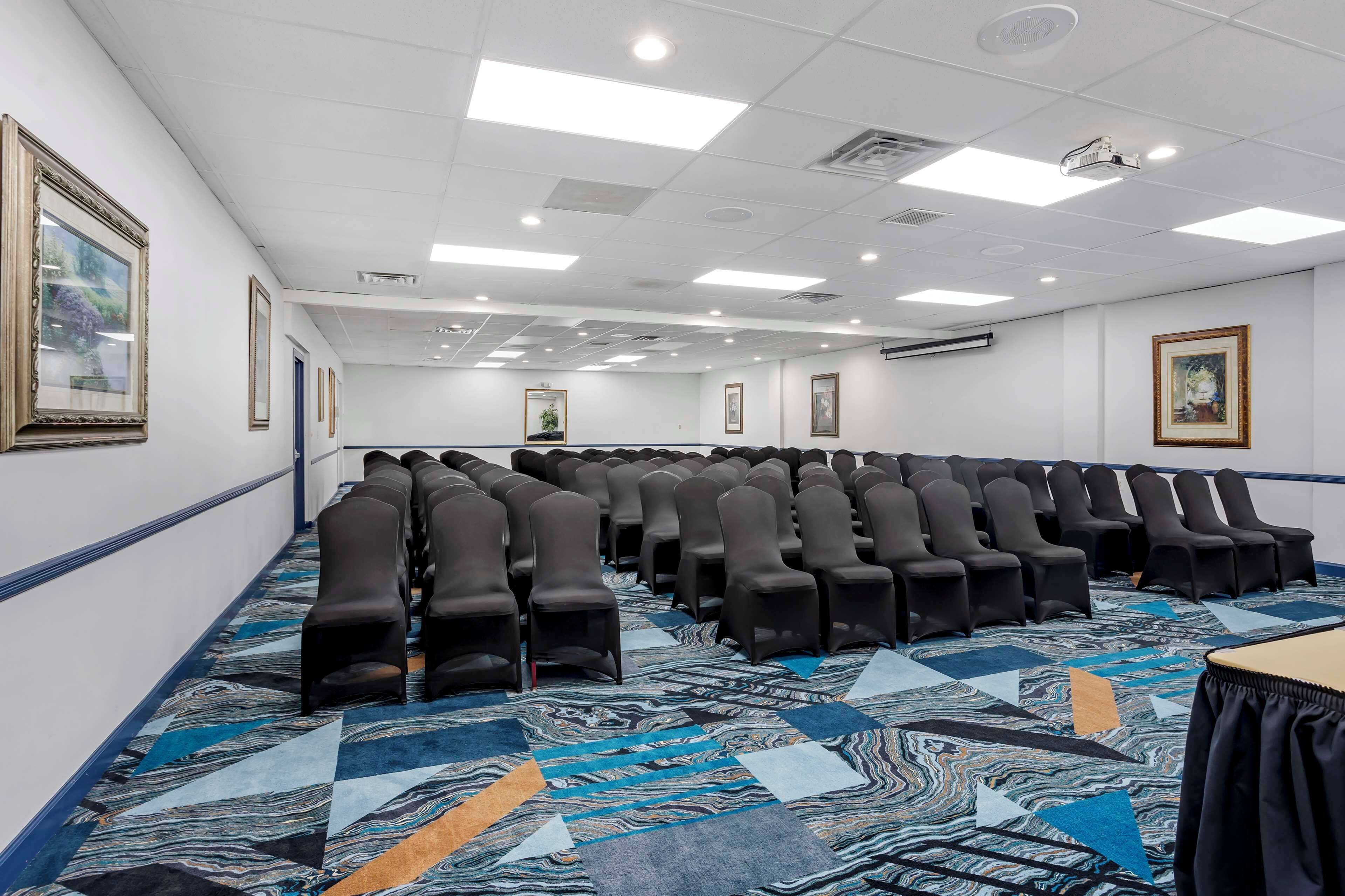 Uma sala de conferências com fileiras de cadeiras pretas, um carpete estampado e obras de arte emolduradas nas paredes.