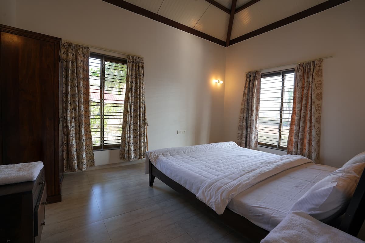 Quarto com duas camas, piso de madeira, guarda-roupa e janelas com cortinas estampadas.