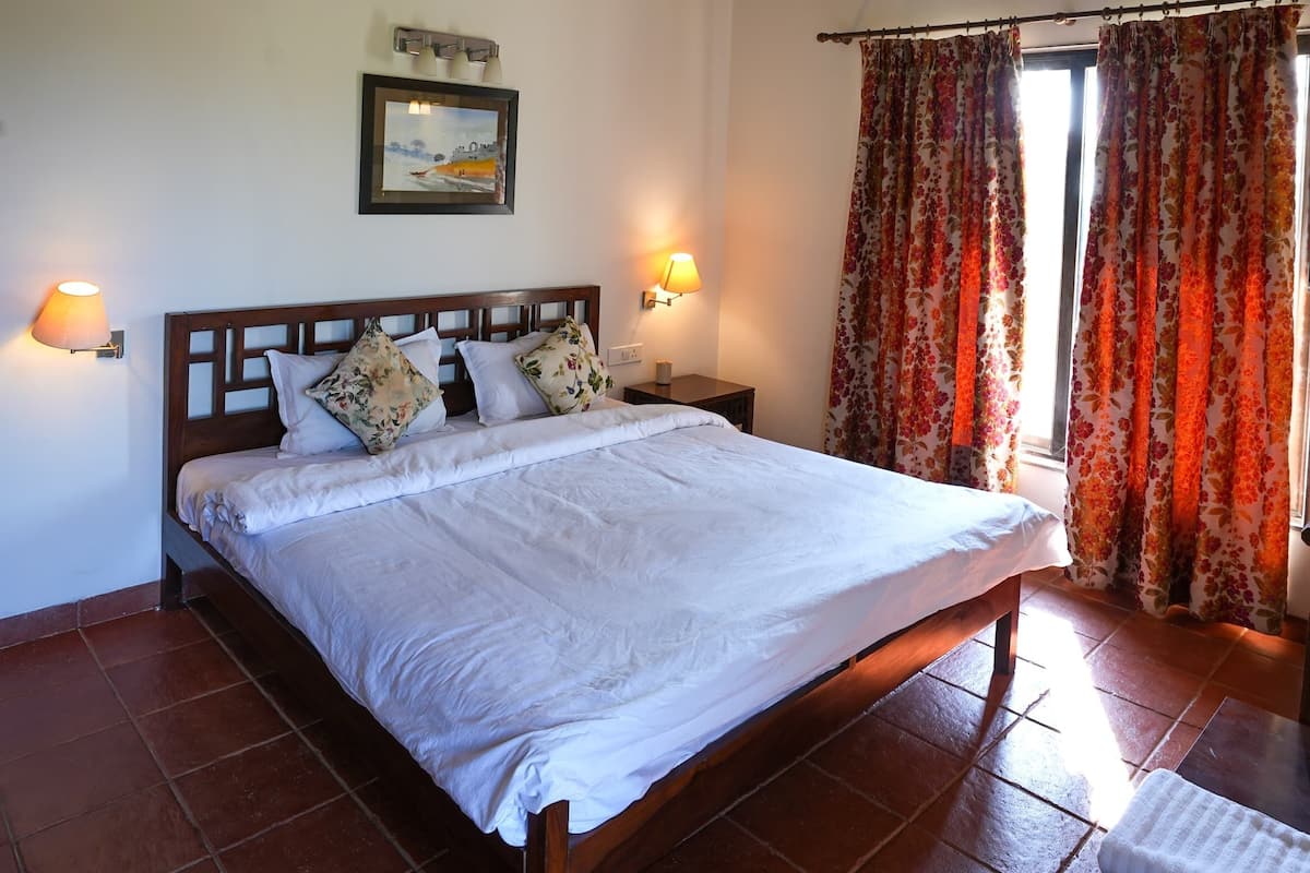 Quarto com cama de madeira, ventilador de teto, mesa de cabeceira com abajur e janela com cortinas.