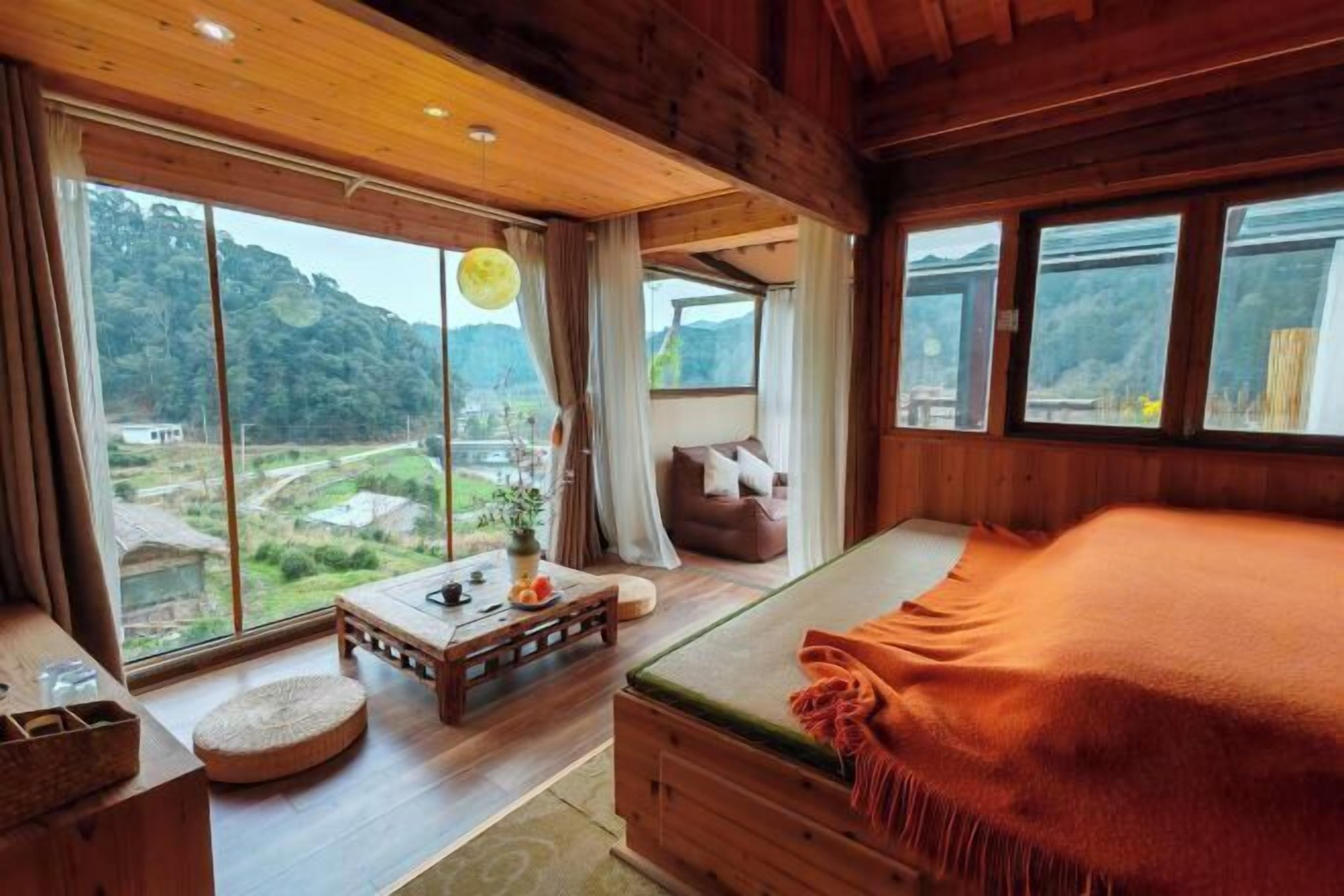 Quarto de cabana de madeira com cama, uma mesinha, uma cadeira e grandes janelas com vista para uma área arborizada.