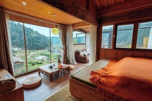 Quarto de cabana de madeira com cama, uma mesinha, uma cadeira e grandes janelas com vista para uma área arborizada.