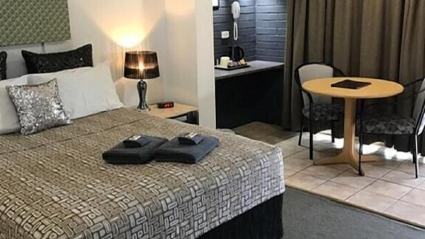 Quarto de hotel com uma cama, escrivaninha com cadeira, ventoinha no teto e telefone.