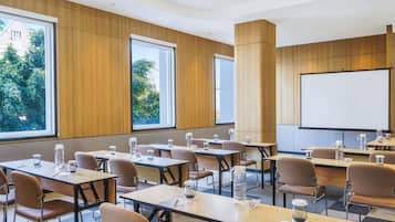 Sala de conferências com mesas, cadeiras, tela de projetor e grandes janelas com vista para árvores.