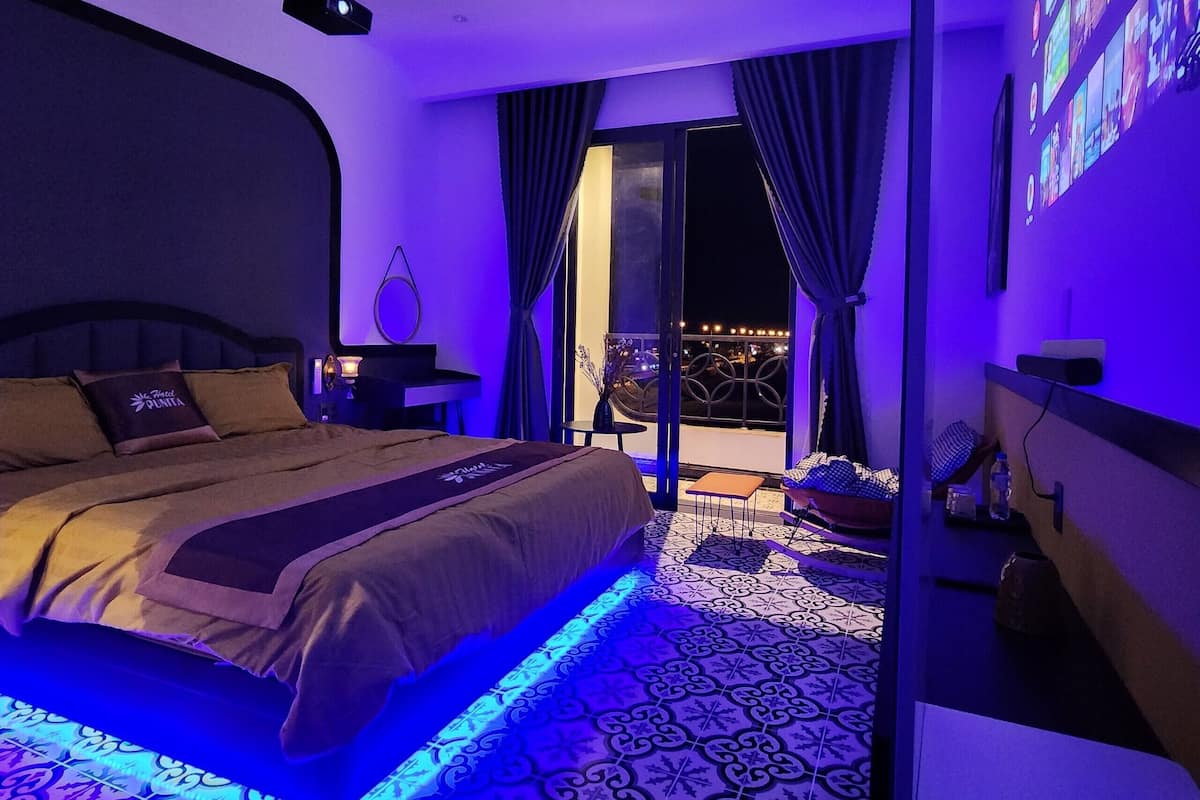 Quarto moderno com cama, mesa de cabeceira, projetor fixado na parede e varanda com guarda-corpo.