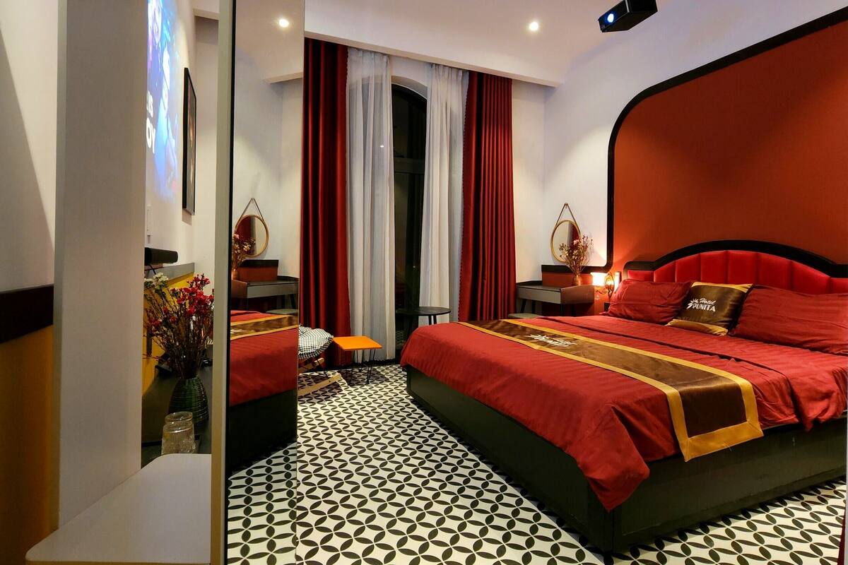 Quarto de hotel moderno com uma cama grande, cortinas vermelhas, uma pequena mesa com um vaso e um chão com padrão geométrico.