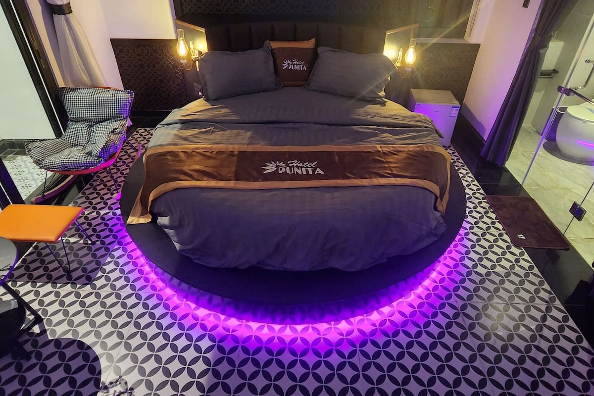 Quarto de hotel com cama, uma cadeira, mesa de cabeceira e banheiro visível por uma porta de vidro.
