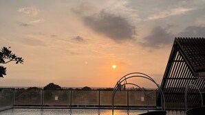 Uma área de piscina no terraço com vista para o pôr do sol sobre o oceano, rodeada por um deck de madeira e espreguiçadeiras.