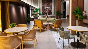 Um hall espaçoso com área de estar central, com cadeiras de vime e uma mesa redonda, cercado por plantas em vasos e um tapete estampado.