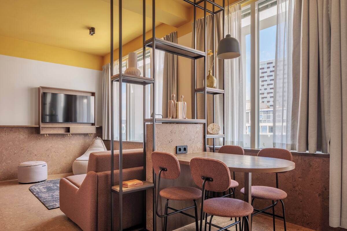 Uma sala de estar moderna com um sofá, uma mesa de jantar com cadeiras, uma TV na parede e uma grande janela com cortinas.