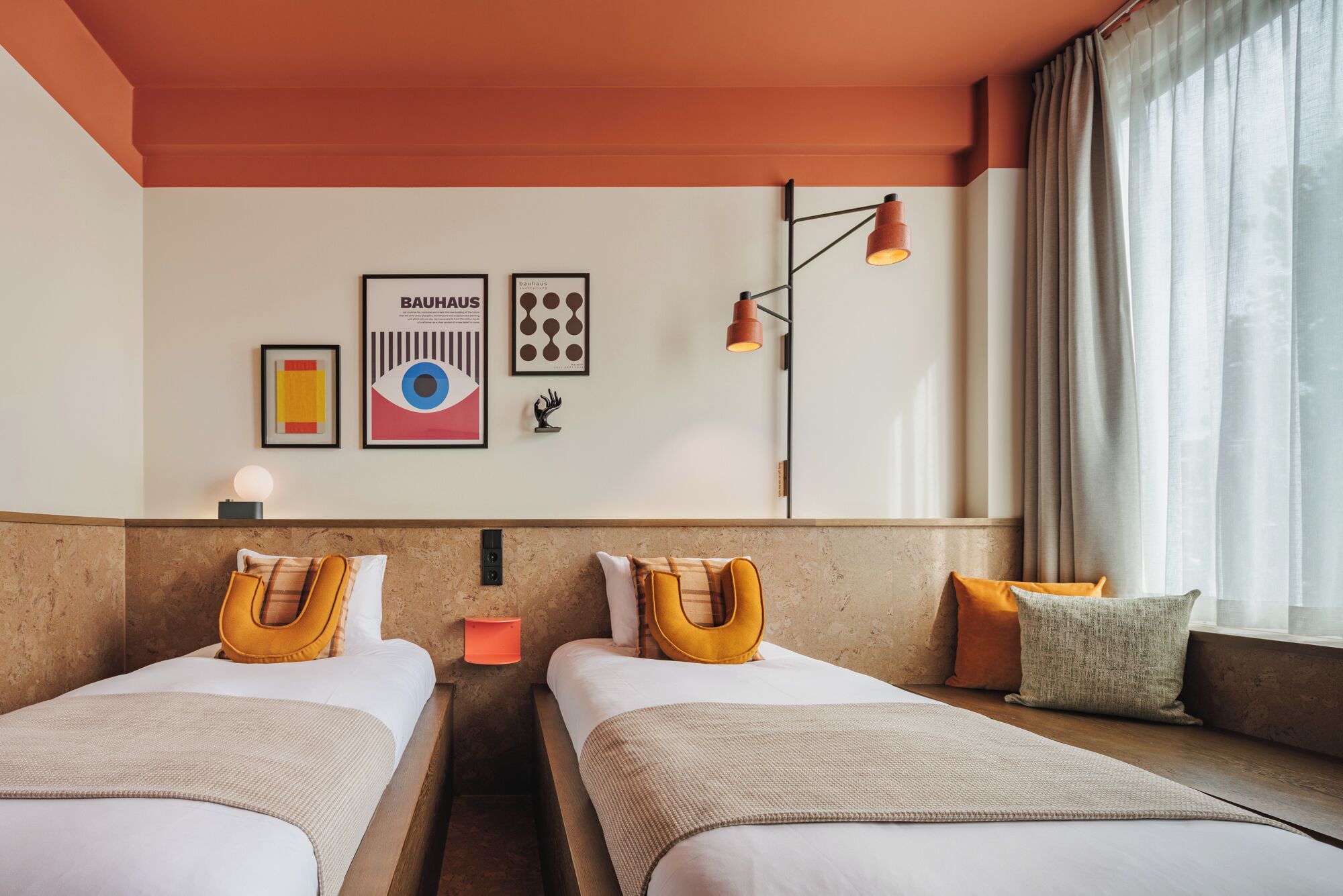 Quarto de hotel com duas camas, uma mesinha de cabeceira, abajur e obras de arte na parede.