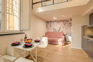 Uma sala de jantar com mesa posta para dois, um sofá rosa e uma área de cozinha com fogão.