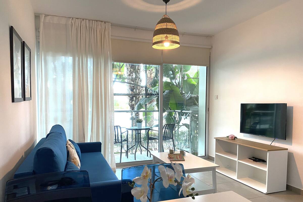 Uma sala de estar moderna com um sofá azul, uma mesa de centro, uma TV em um suporte e uma varanda com plantas.