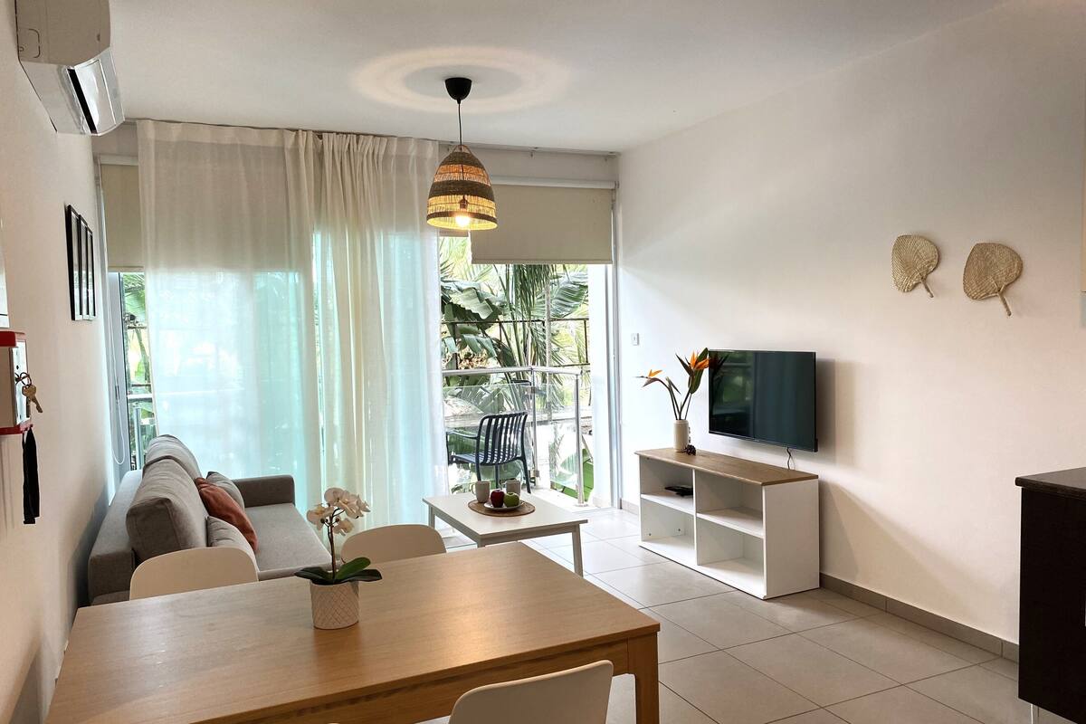Uma sala de estar moderna com sofá, mesa de jantar, cadeiras, uma TV em um suporte de madeira e uma varanda com vista para o verde.