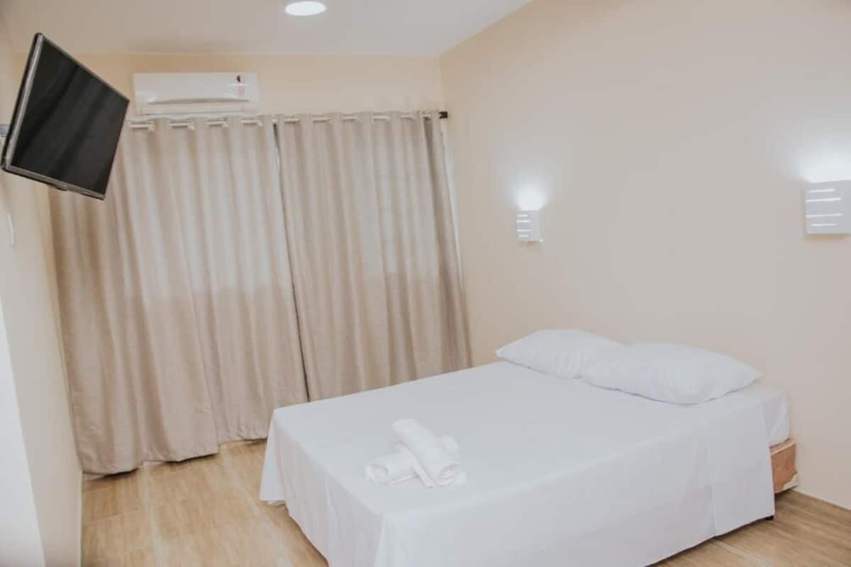 Uma cama de solteiro com roupa de cama branca, uma TV de tela plana fixada na parede, uma janela com cortinas bege e um aparelho de luz fixado na parede.
